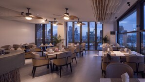 Restaurante moderno com pé-direito alto, ventiladores de teto e grandes janelas com vista para a cidade.