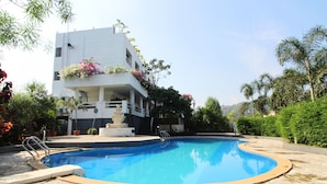 Piscina com deck de pedra, um edifício branco com varandas e vegetação ao redor.