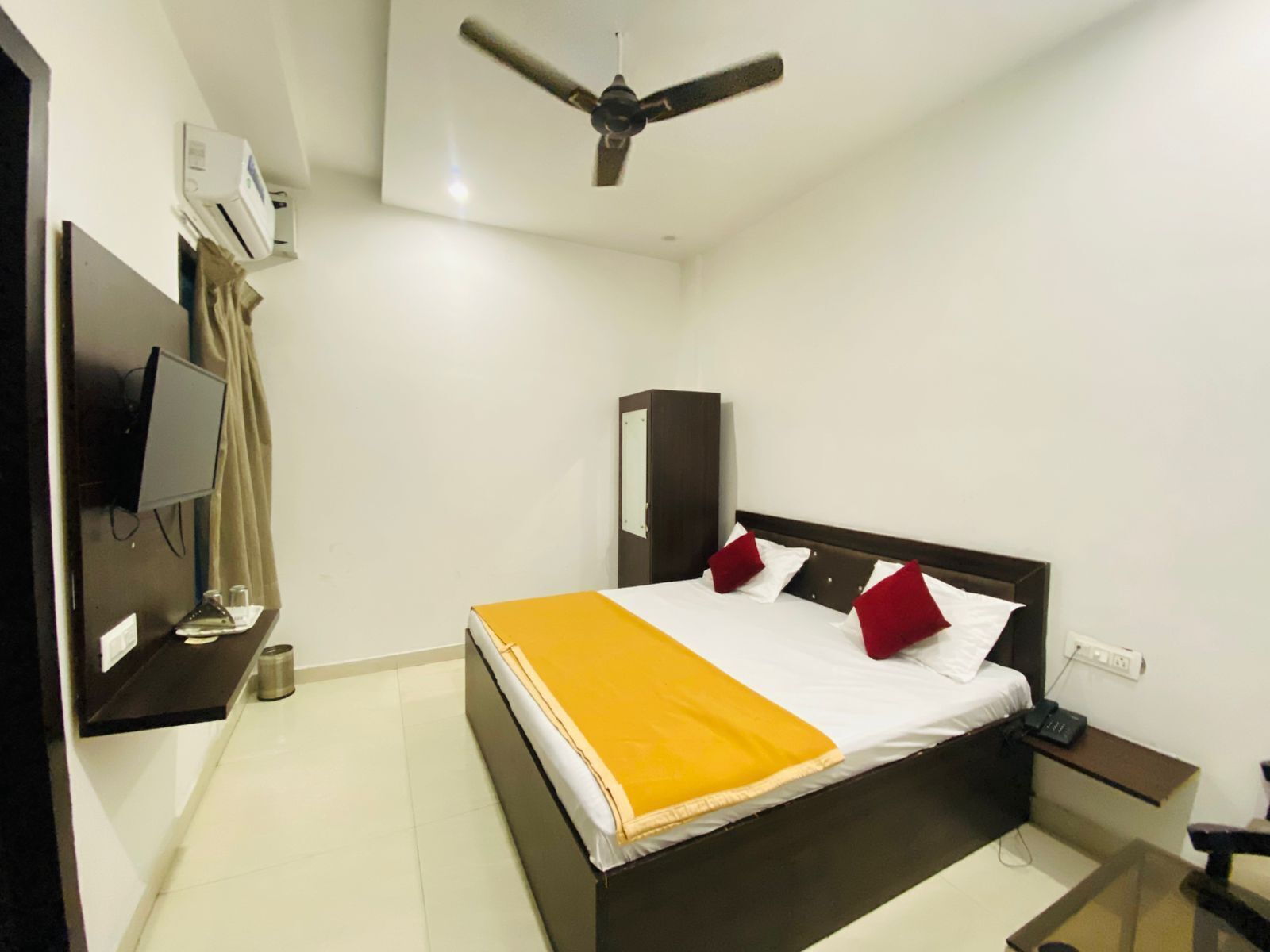 Quarto de hotel com cama, ventilador de teto, televisão, minibar e mesa de cabeceira.