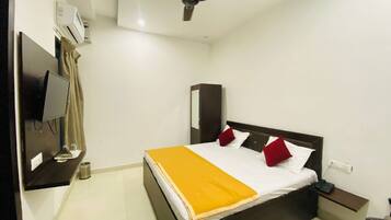 Quarto de hotel com cama, ventilador de teto, televisão, minibar e mesa de cabeceira.