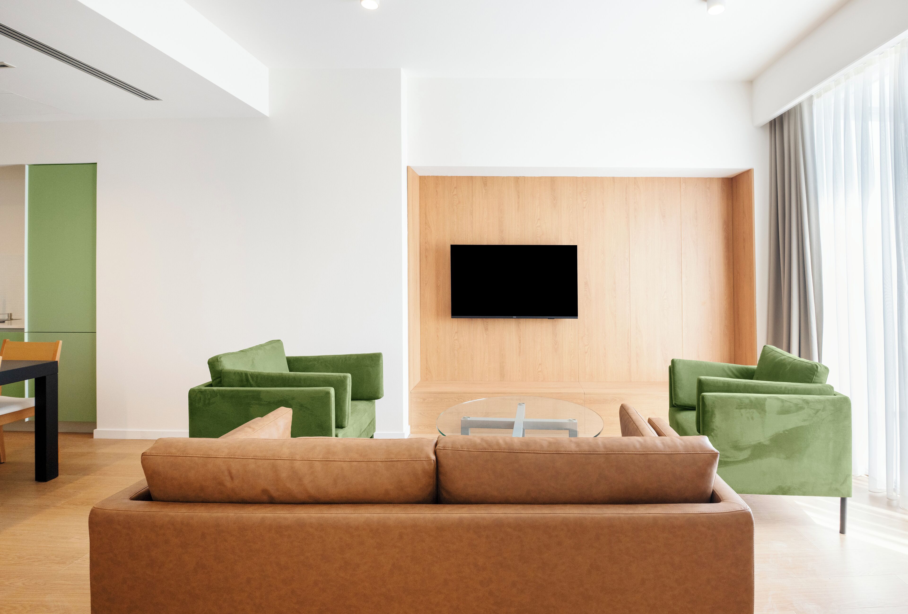 Uma sala de estar moderna com um sofá marrom, poltronas verdes, uma mesa de centro de vidro e uma TV de tela plana fixada numa parede de madeira.