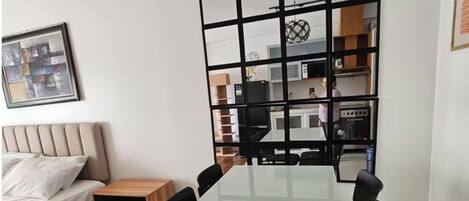Quarto com mesa de jantar de vidro, sofá, quadro na parede e área de cozinha visível através de uma divisória preta.