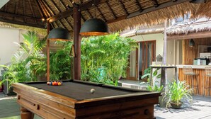 Mesa de bilhar em ambiente tropical, com telhado de palha e vegetação exuberante.