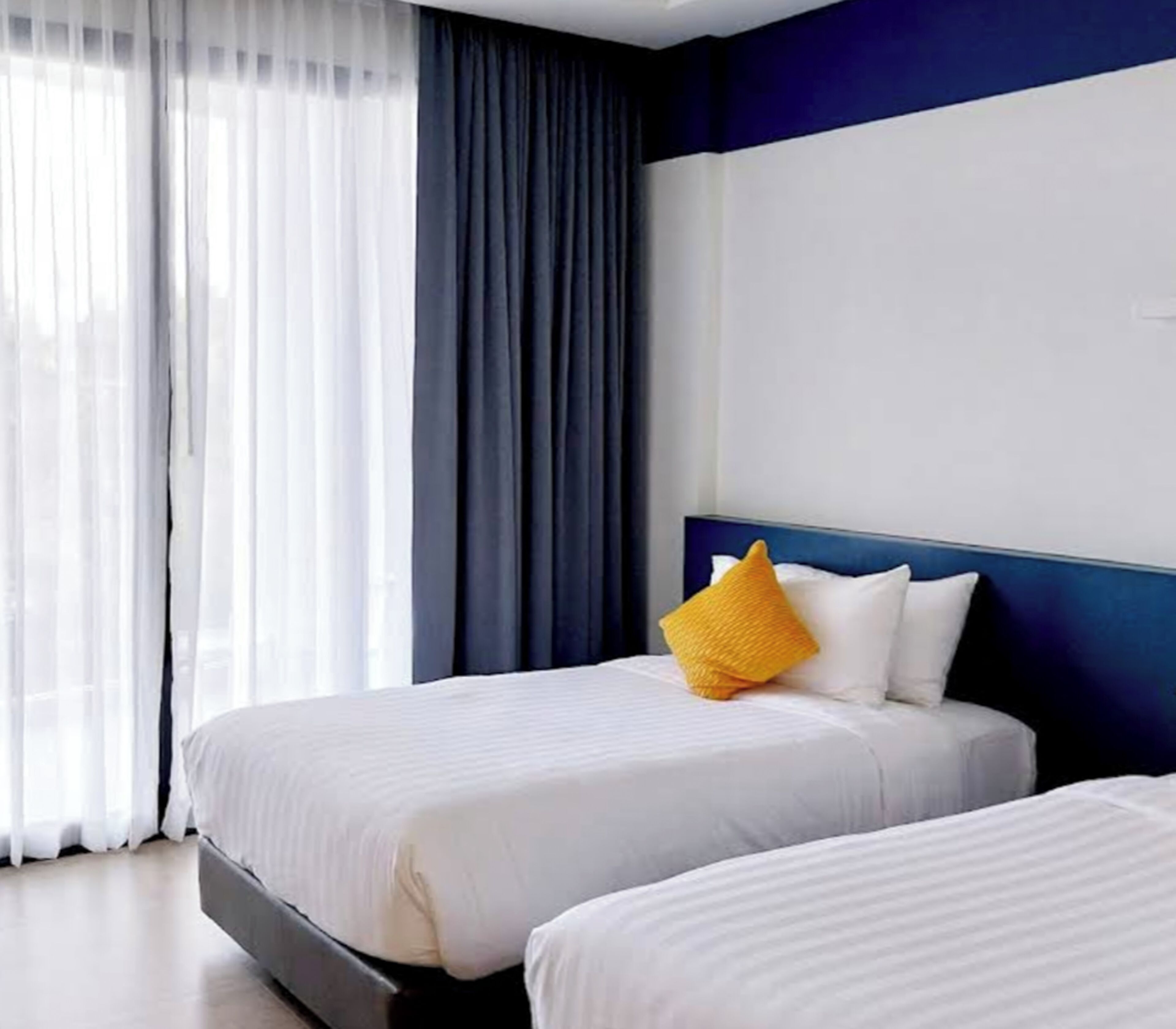 Quarto de hotel com duas camas, uma janela grande com cortinas finas e uma parede de destaque azul escura.