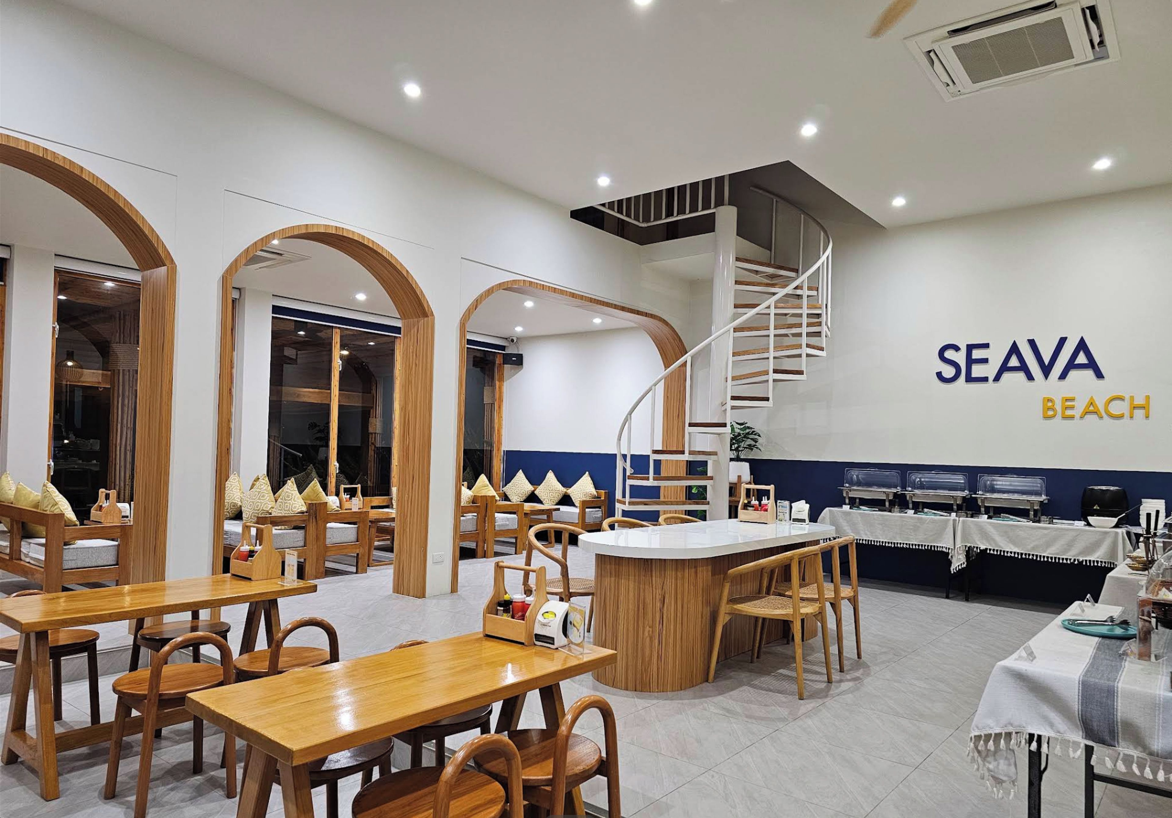 Interior de restaurante com mesas e cadeiras de madeira, uma ilha central e uma escadaria que leva a um andar superior.