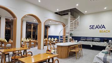 Interior de restaurante com mesas e cadeiras de madeira, uma ilha central e uma escadaria que leva a um andar superior.