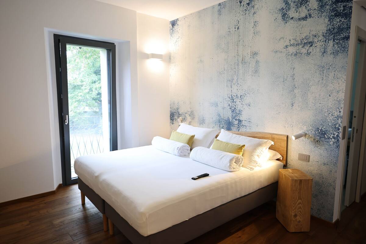 Quarto com cama, mesa de cabeceira de madeira, luminária fixada na parede e porta que dá para uma varanda.