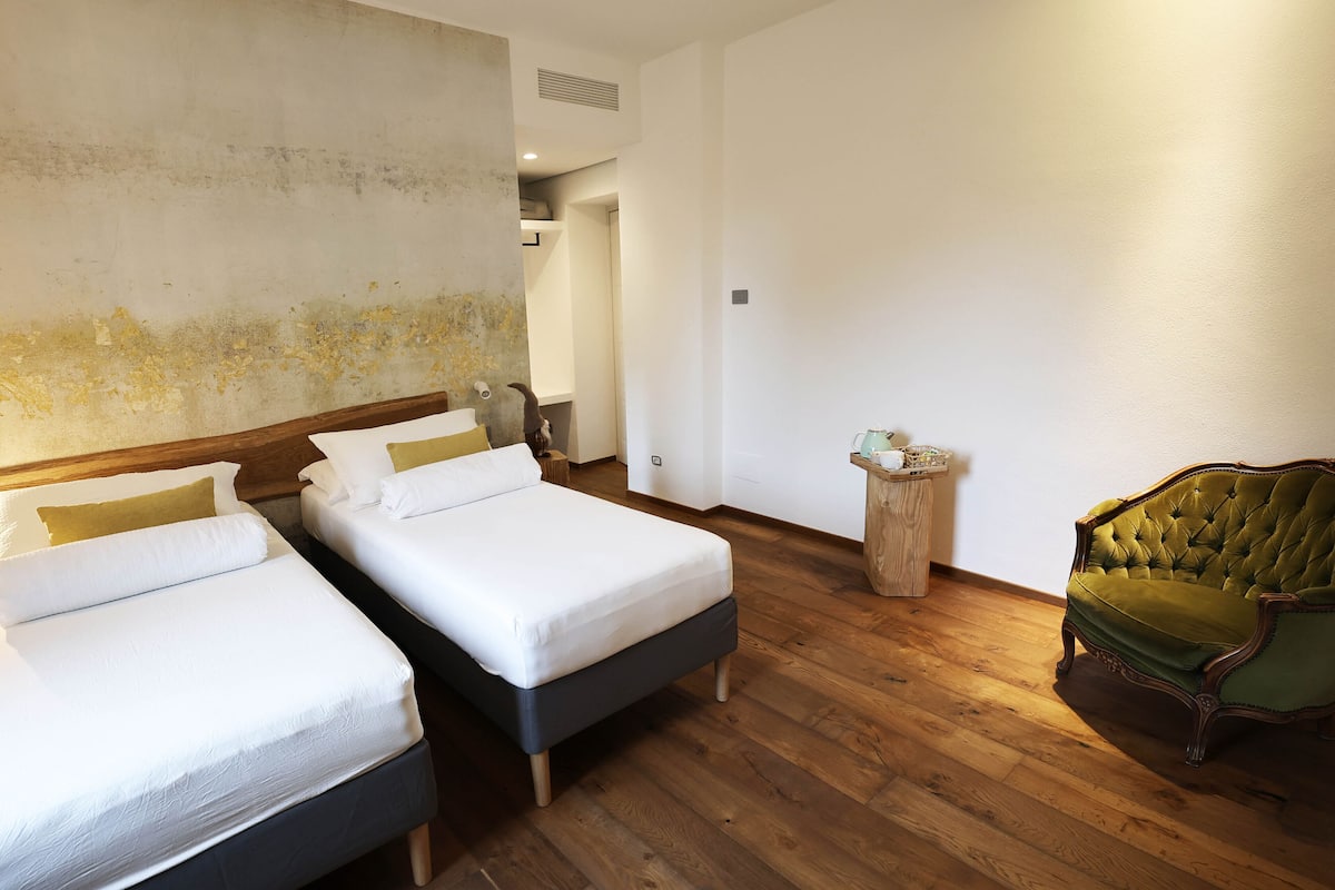 Quarto de hotel com duas camas, piso de madeira, uma poltrona verde e uma pequena mesa de apoio de madeira com um chaleira.
