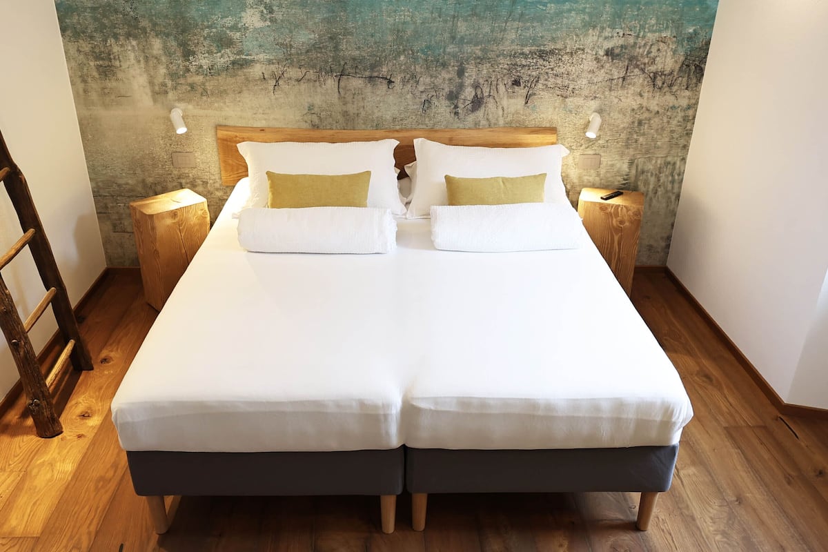 Cama de casal com lençóis brancos e duas almofadas, cabeceira de madeira, mesas de cabeceira com luminárias e uma parede rústica com mural texturizado.