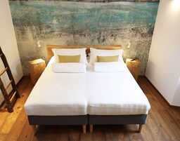 Cama de casal com lençóis brancos e duas almofadas, cabeceira de madeira, mesas de cabeceira com luminárias e uma parede rústica com mural texturizado.