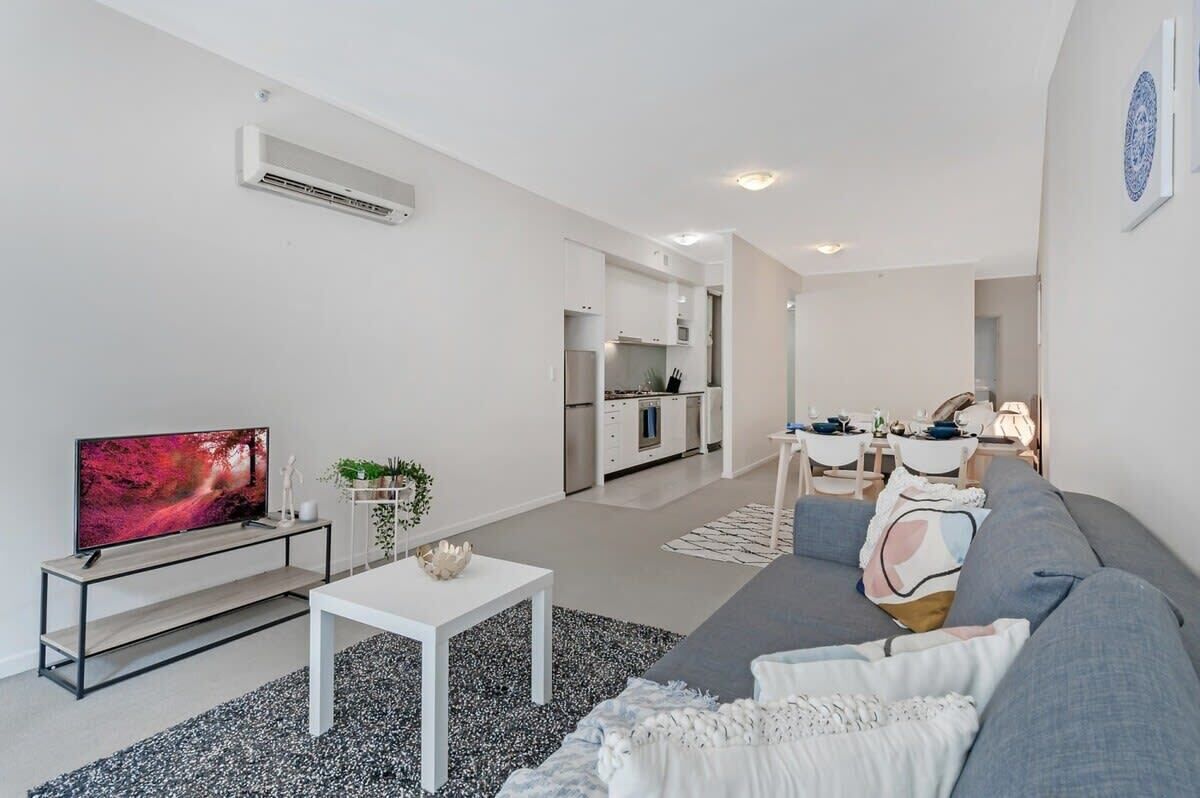 Uma sala de estar moderna com um sofá cinza, uma TV de tela plana, uma pequena mesa branca e uma cozinha ao fundo.