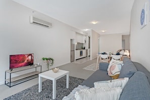 Uma sala de estar moderna com um sofá cinza, uma TV de tela plana, uma pequena mesa branca e uma cozinha ao fundo.