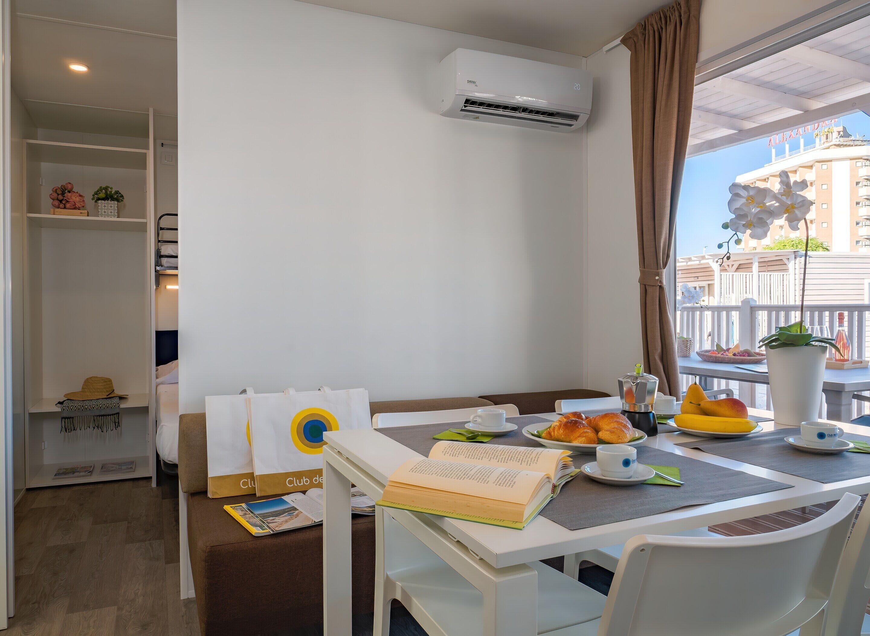Quarto de hotel com mesa de jantar posta para o café da manhã, janela com vista para um edifício e aparelho de ar condicionado.