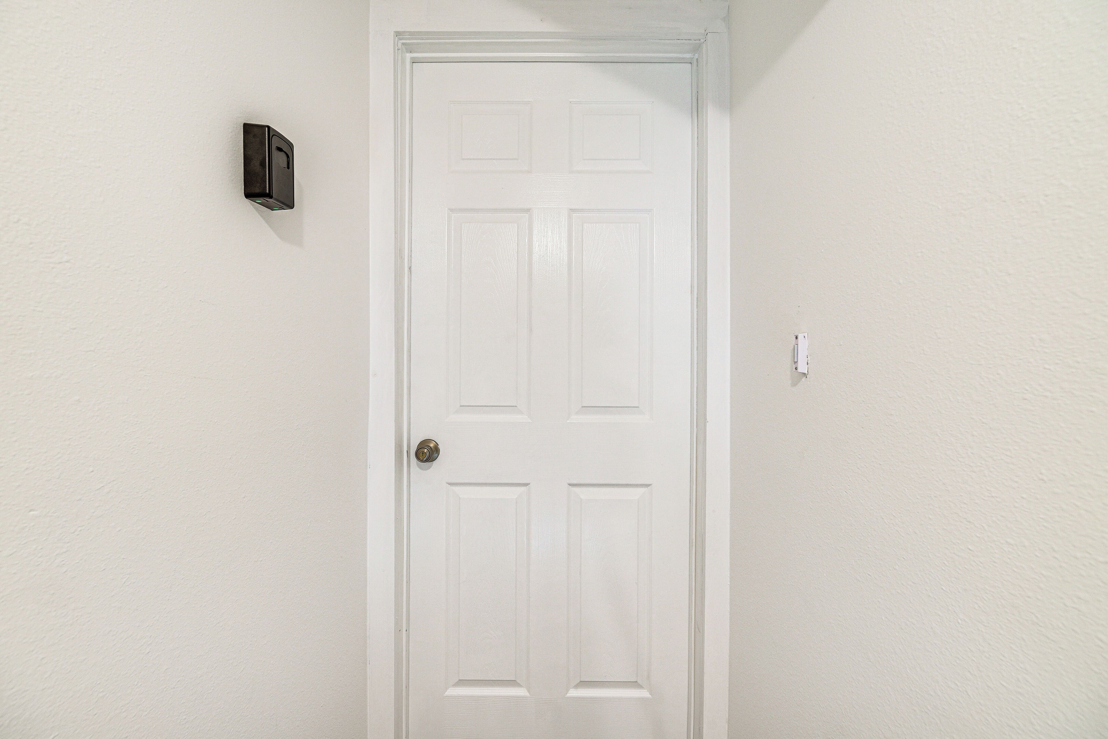 Uma porta branca com design simples, um pequeno dispositivo fixado na parede à esquerda e um interruptor de luz à direita.