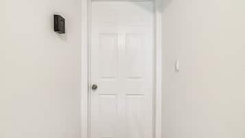 Uma porta branca com design simples, um pequeno dispositivo fixado na parede à esquerda e um interruptor de luz à direita.