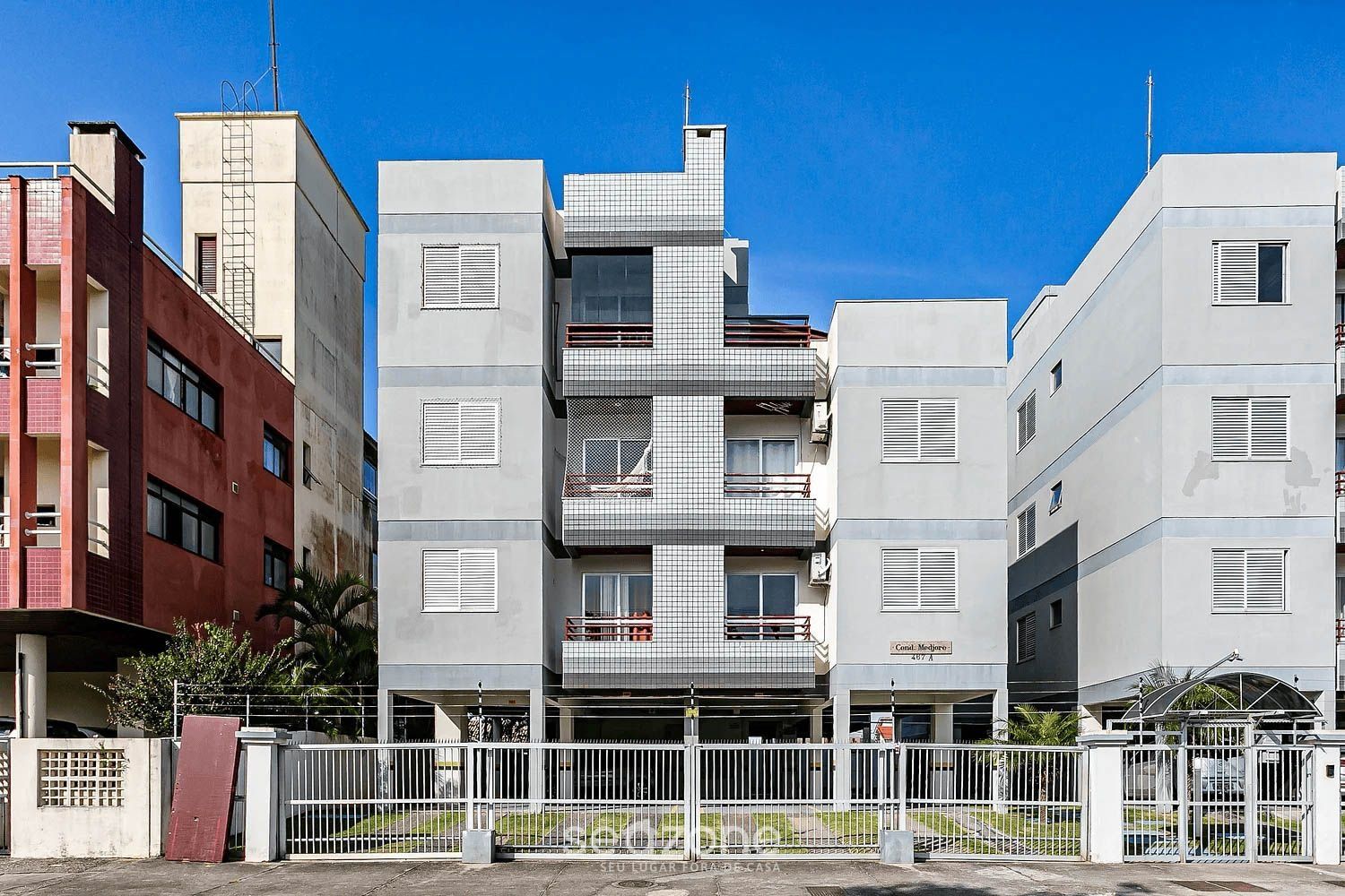 Edifício residencial com vários andares, varandas, portão fechado e céu claro.