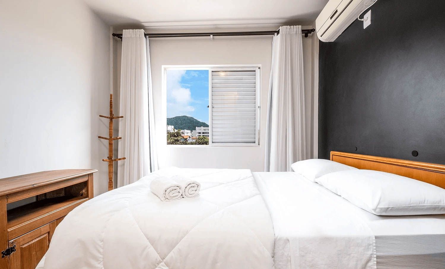 Quarto de hotel com cama, mesa de cabeceira de madeira, janela com cortinas e vista para edifícios e uma colina.