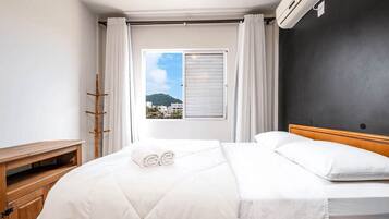 Quarto de hotel com cama, mesa de cabeceira de madeira, janela com cortinas e vista para edifícios e uma colina.