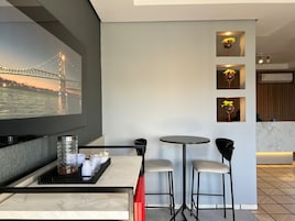 Quarto moderno com uma TV grande exibindo uma vista da cidade, uma pequena área de refeições com uma mesa e dois banquetes, e uma prateleira fixada na parede com objetos decorativos.
