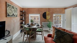 Uma sala de jantar com uma mesa redonda, cadeiras e uma televisão sobre um suporte. Há uma estante de livros e um vaso com planta.