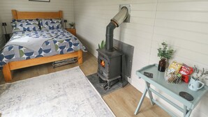 Quarto com cama, cabeceira de madeira, mesinha com um vaso e petiscos, e lareira a lenha.
