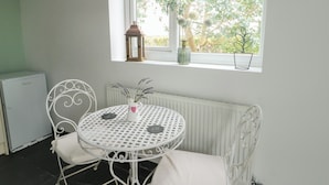 Um quarto pequeno com um radiador branco, uma mesa redonda com duas cadeiras, uma janela com vista para árvores e um pequeno candeeiro no parapeito.