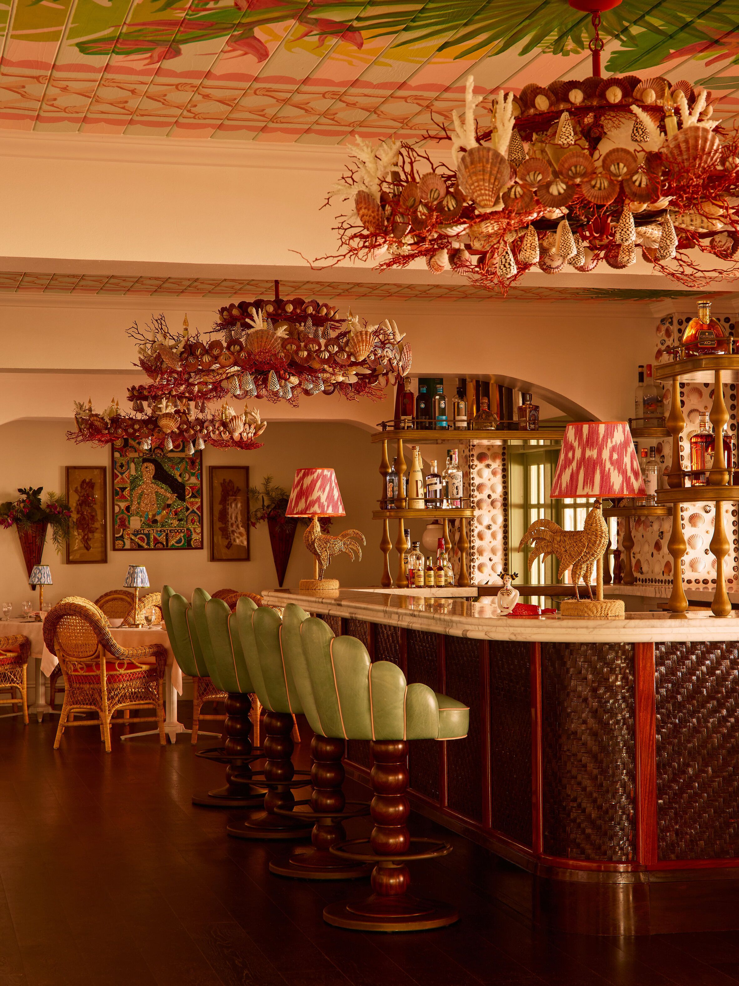 Uma área de bar com teto alto, conchas decorativas e diversos tipos de garrafas. Há banquetas de bar estofadas em verde e uma mesa com abajur.