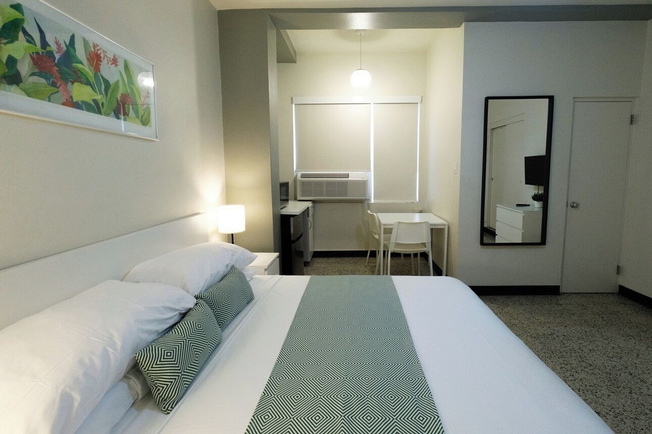 Quarto de hotel com uma cama, uma escrivaninha, uma cadeira, um espelho e uma janela.