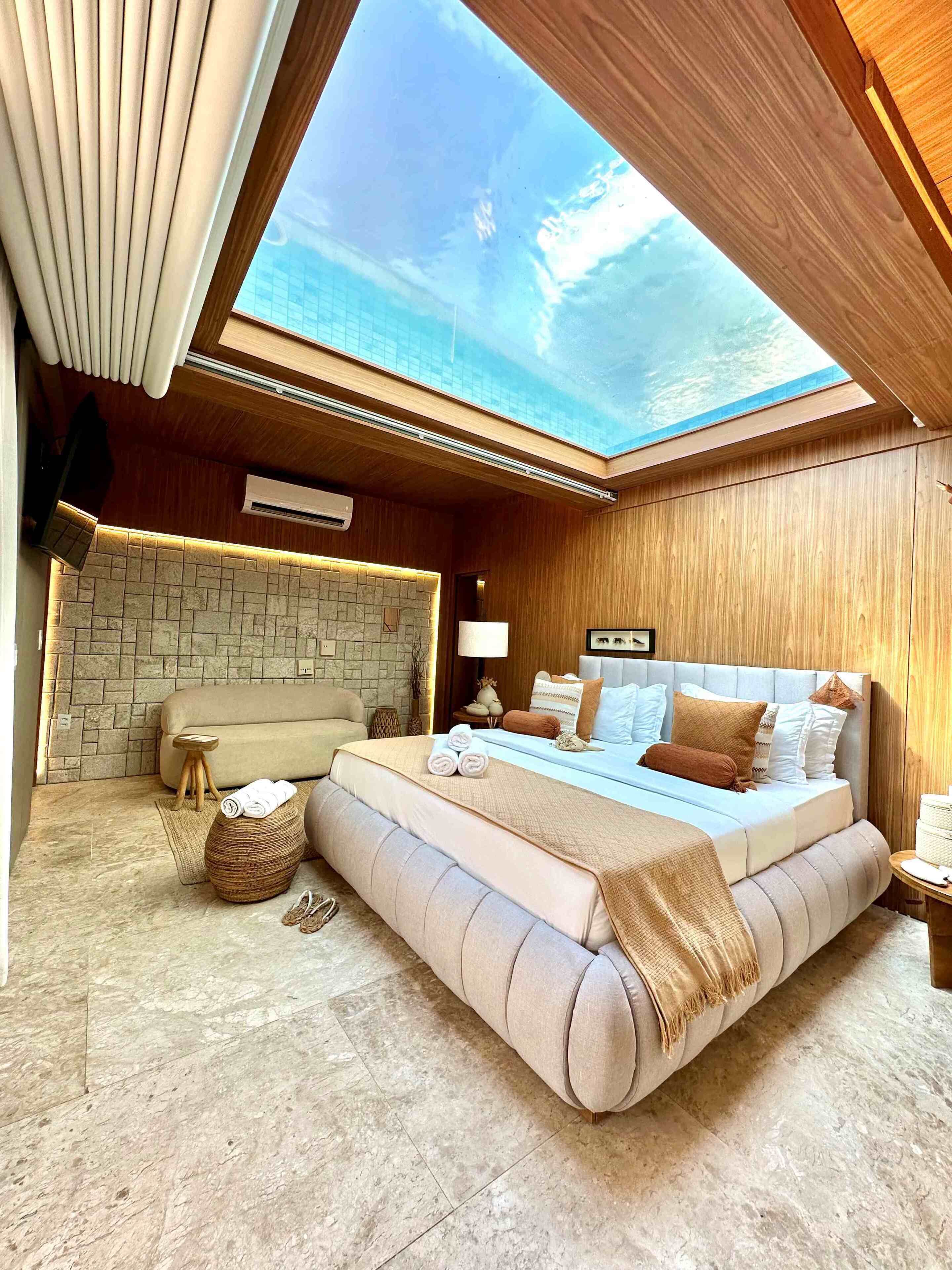 Quarto com claraboia, parede de pedra, cama e um pequeno banquinho de madeira.