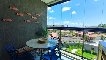 Varanda com mesa redonda, cadeiras azuis, parede decorativa com formas de peixe e vista para edifícios e vegetação.