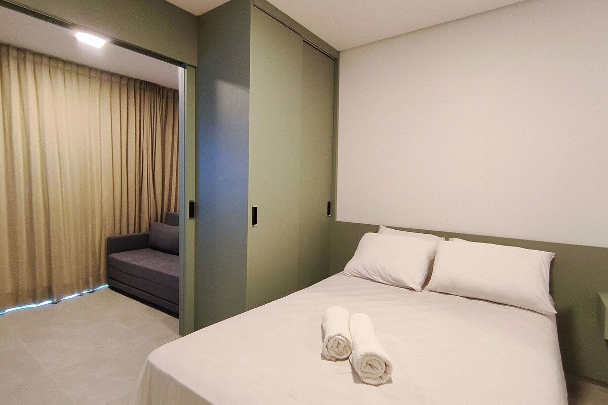 Quarto de hotel com cama, duas almofadas, mesa de cabeceira, cadeira e cortina.