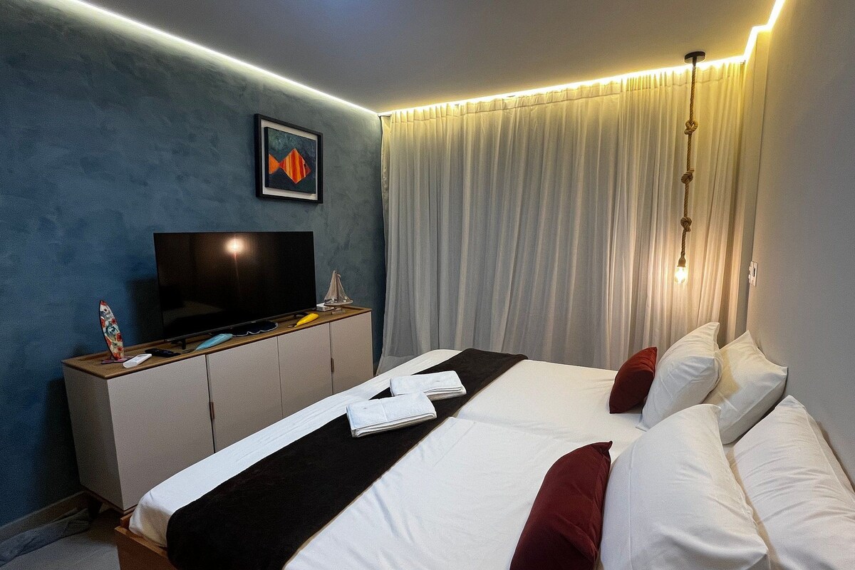 Quarto de hotel com cama, televisão, minibar e cortina.