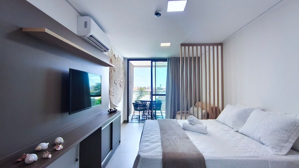 Quarto de hotel moderno com televisão de tela plana, uma cama, varanda com mesa e cadeiras, e vista para a paisagem urbana.