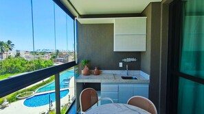 Uma varanda com vista para uma piscina, uma pequena cozinha com pia e armários, e duas cadeiras de vime.