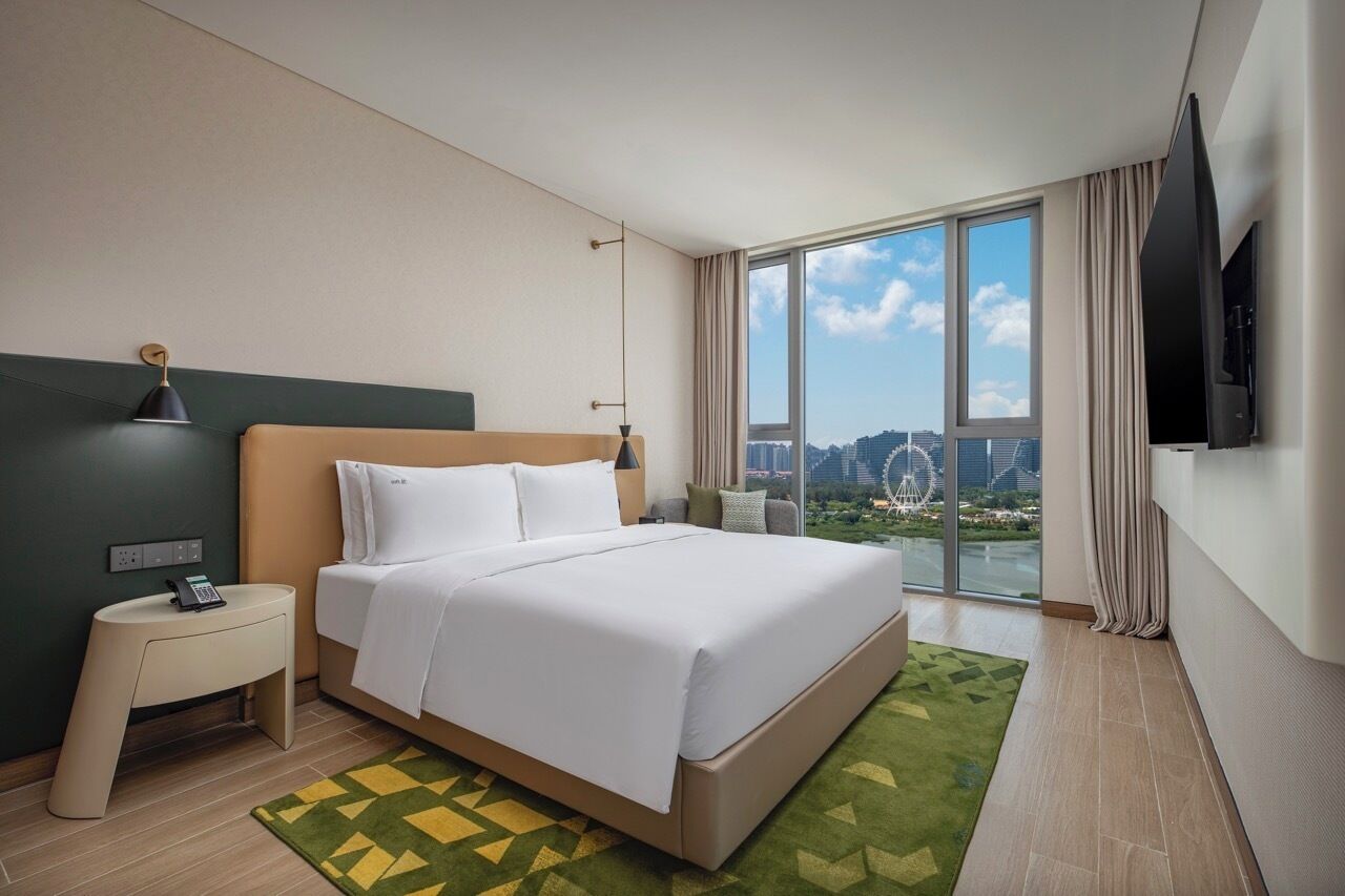 Quarto de hotel com cama, mesa de cabeceira, telefone, televisão e uma grande janela com vista para a cidade.