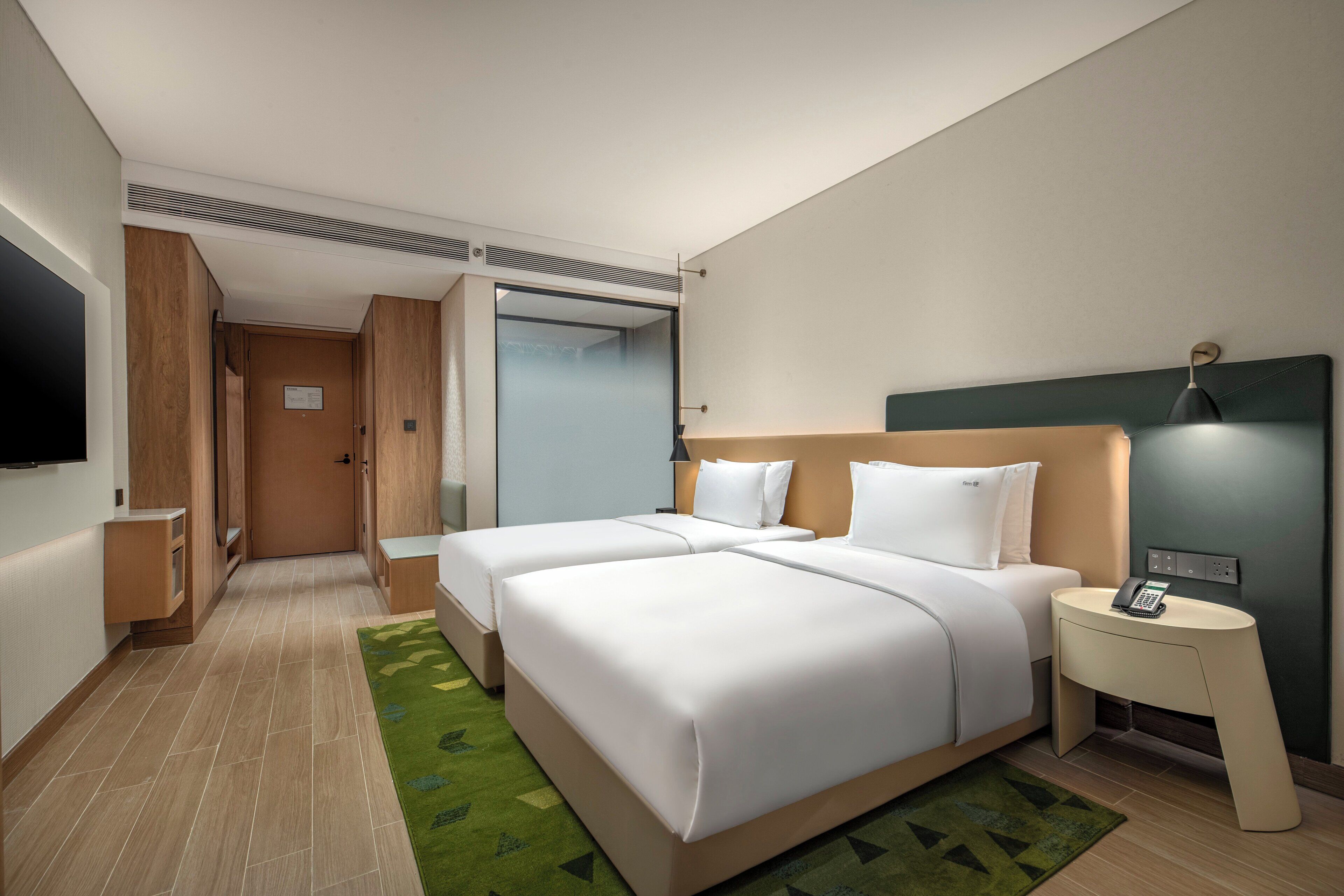 Quarto de hotel com duas camas, uma televisão de tela plana na parede, mesa de cabeceira com abajur e tapete verde.