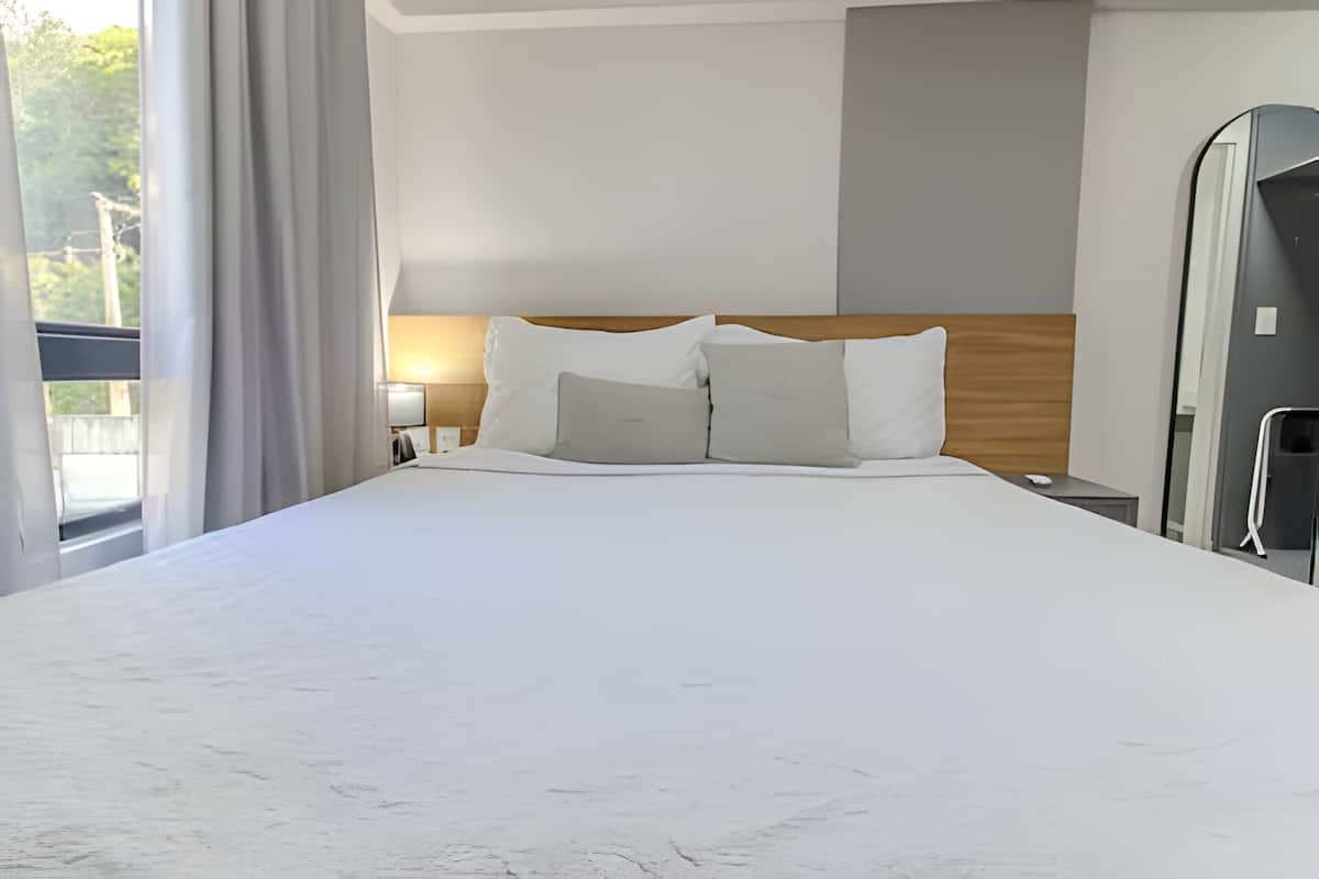 Quarto de hotel com uma cama grande, cabeceira de madeira, mesa de cabeceira, abajur e vista para o exterior.