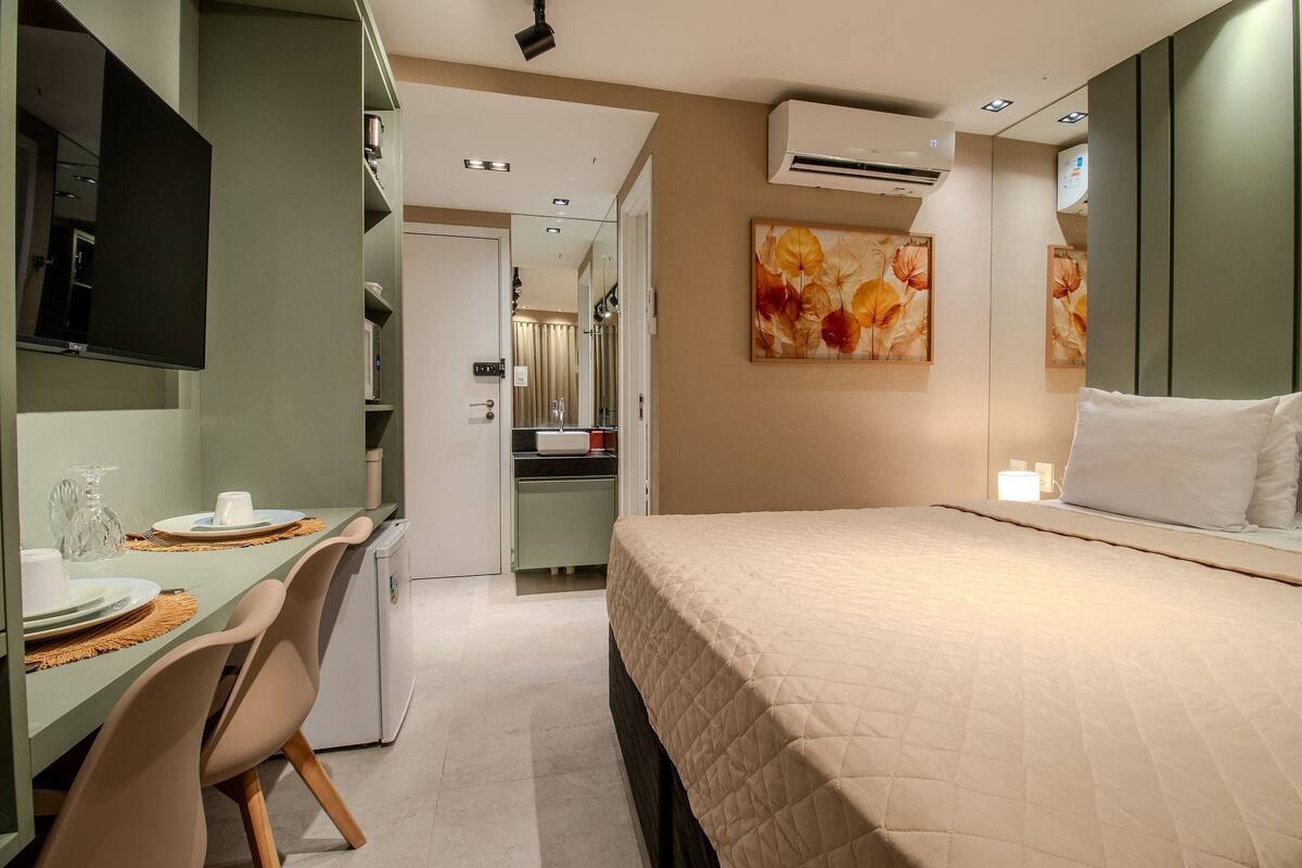 Quarto de hotel com uma cama, uma mesa com cadeiras, um espelho e um quadro na parede.