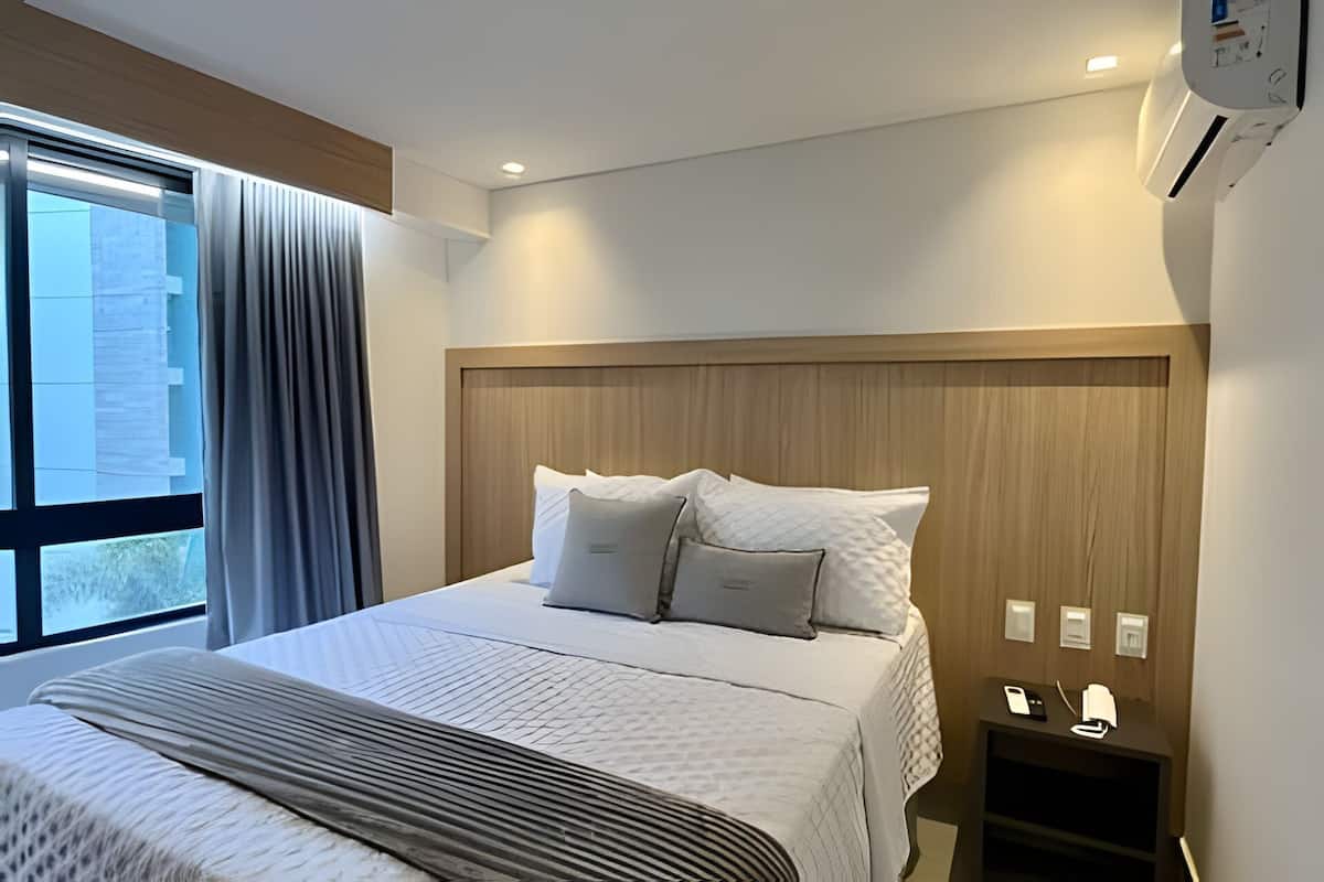 Quarto de hotel com uma cama grande, cabeceira de madeira, mesa de cabeceira com telefone e abajur, e uma janela com cortinas.