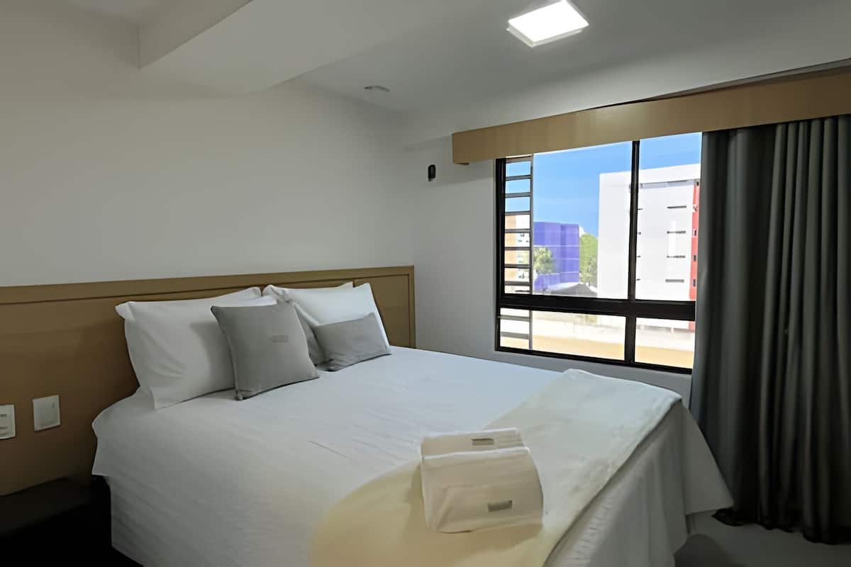 Quarto de hotel com cama, janela com cortinas, luminária no teto e vista para prédios.