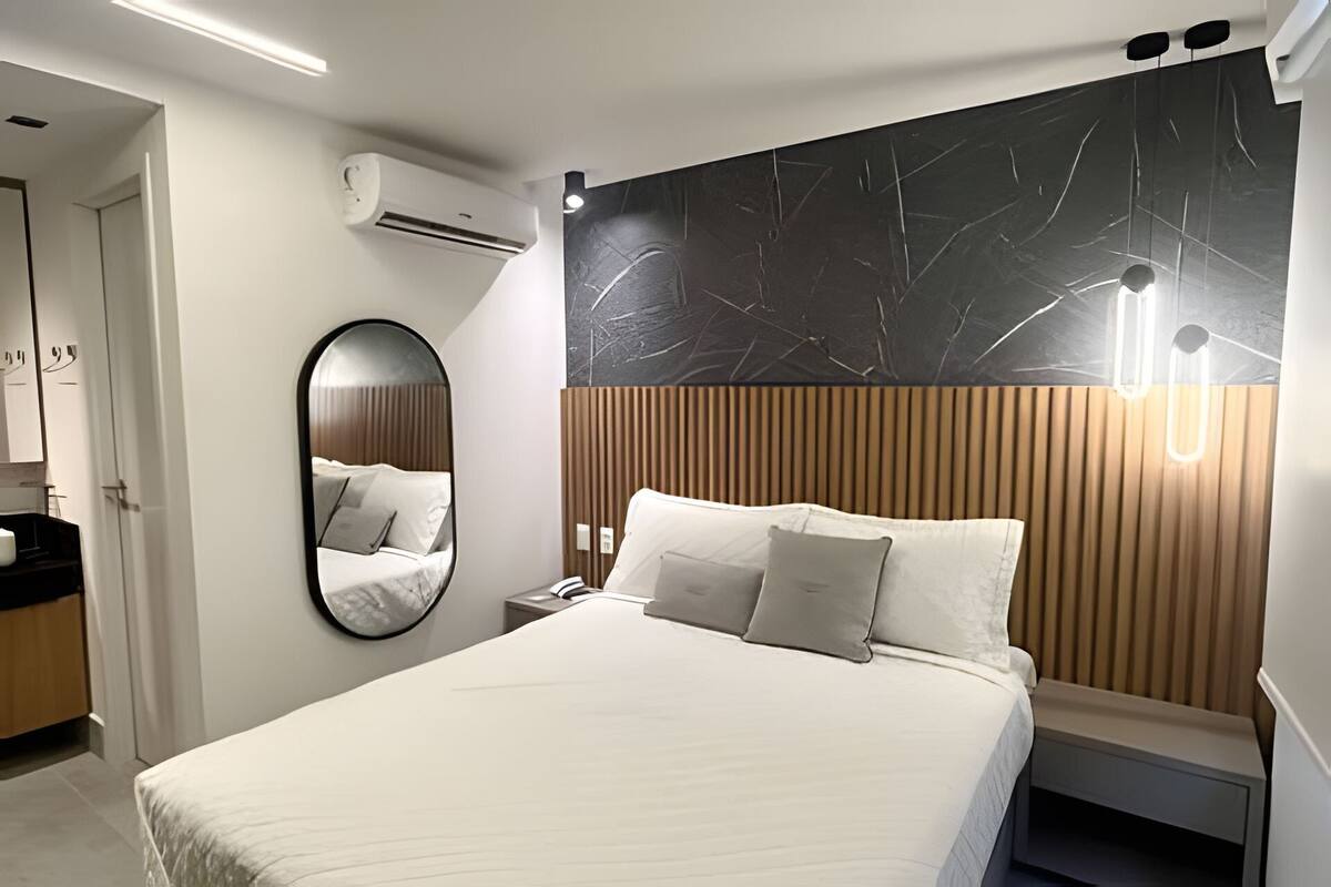 Quarto de hotel moderno com uma cama, um espelho, um aparelho de ar condicionado, uma cabeceira de madeira e uma mesinha de cabeceira.