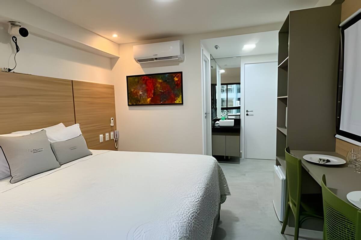 Quarto de hotel com uma cama, uma escrivaninha, uma cadeira, ar condicionado, um quadro e um banheiro visível através de uma porta aberta.