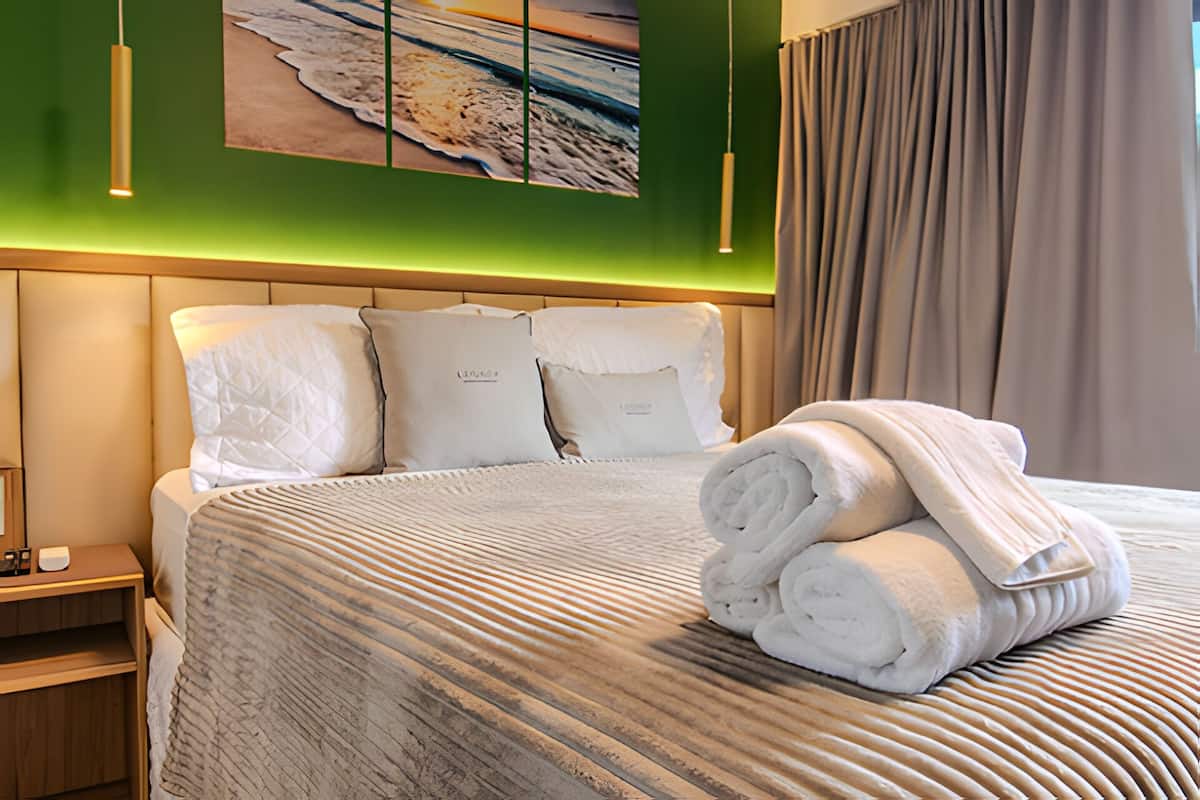 Uma cama bem arrumada com colcha listrada, uma toalha dobrada e um quadro com uma cena de praia na parede.