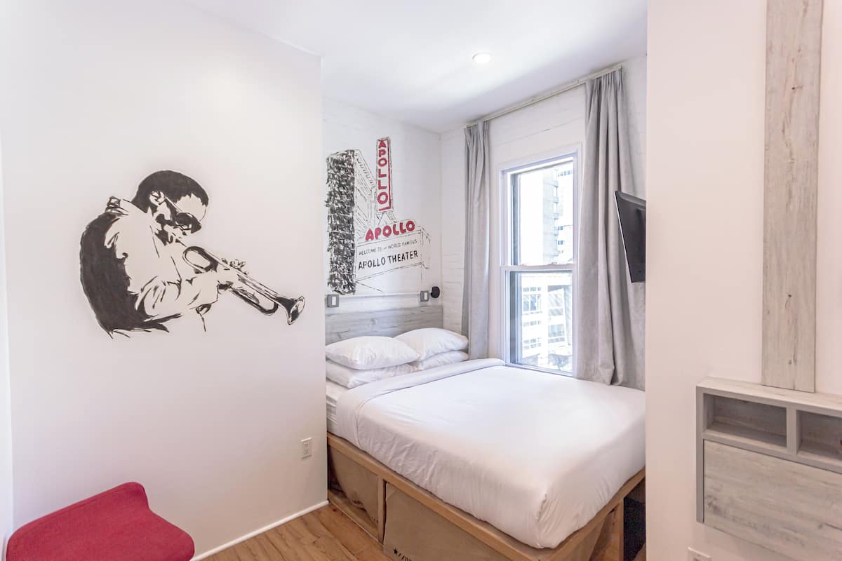 Quarto com cama, cadeira vermelha, painel na parede, janela e televisão.