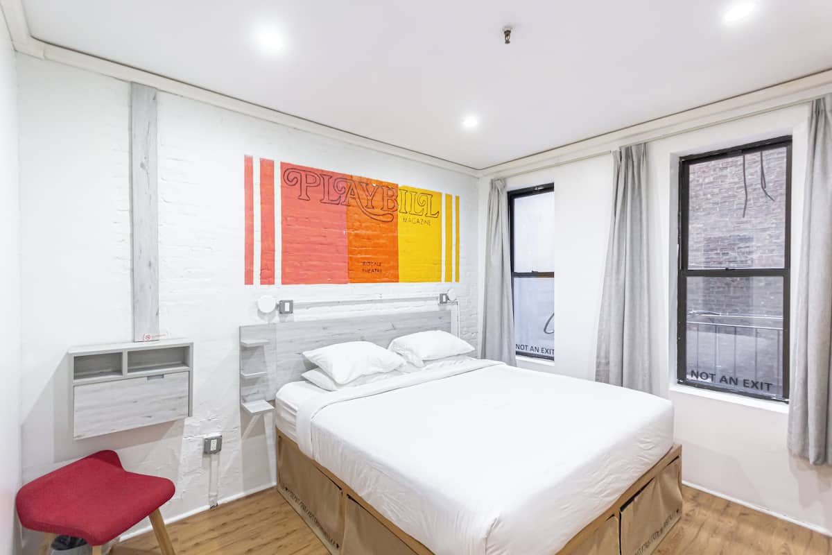 Quarto moderno com cama, uma cadeira vermelha, parede branca com um cartaz da revista Playbill, janela com cortinas e uma placa com a inscrição 'NÃO É SAÍDA'.