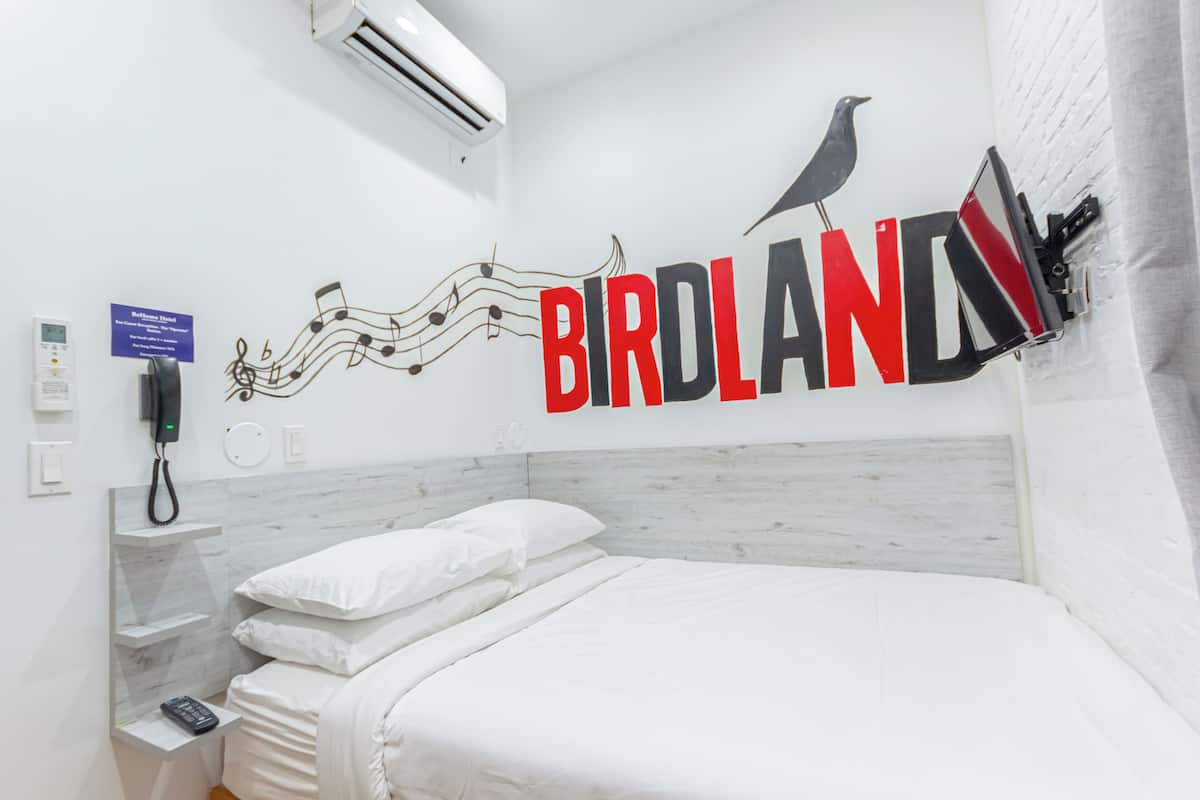 Quarto de hotel com cama, mesa de cabeceira com telefone, ar condicionado fixado na parede e placa indicando o tipo de quarto.