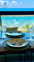 Uma mesa de jantar posta com pratos, copos e talheres, com vista para o oceano através de uma janela.