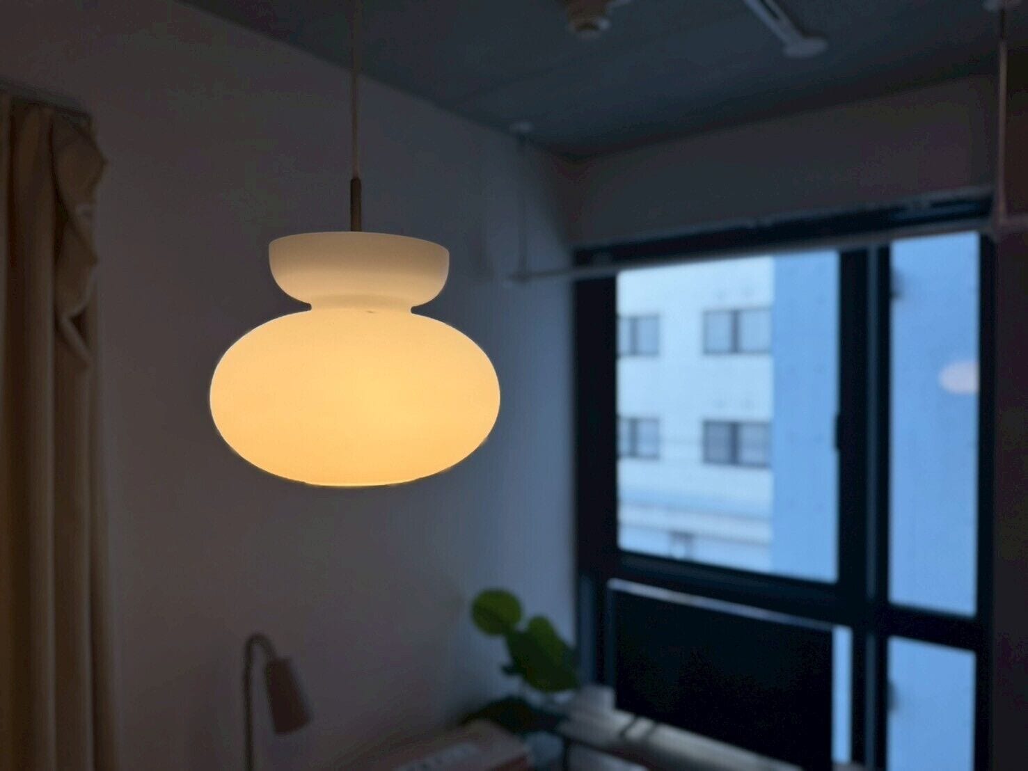 Quarto moderno com luminária pendente, janela com vista para prédios e uma planta no parapeito.
