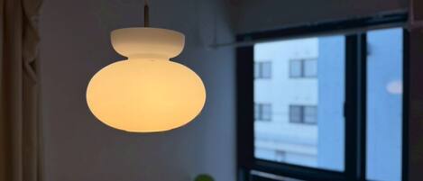 Quarto moderno com luminária pendente, janela com vista para prédios e uma planta no parapeito.