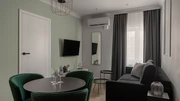 Uma sala de estar moderna com uma mesa redonda, duas cadeiras verdes, um sofá cinza, uma TV de tela plana, um espelho e uma janela com cortinas.